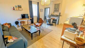 Uma sala de estar com sofá, mesa de centro e poltronas, uma televisão, um espaço de jantar com mesa e cadeiras, e uma cozinha com geladeira e micro-ondas.