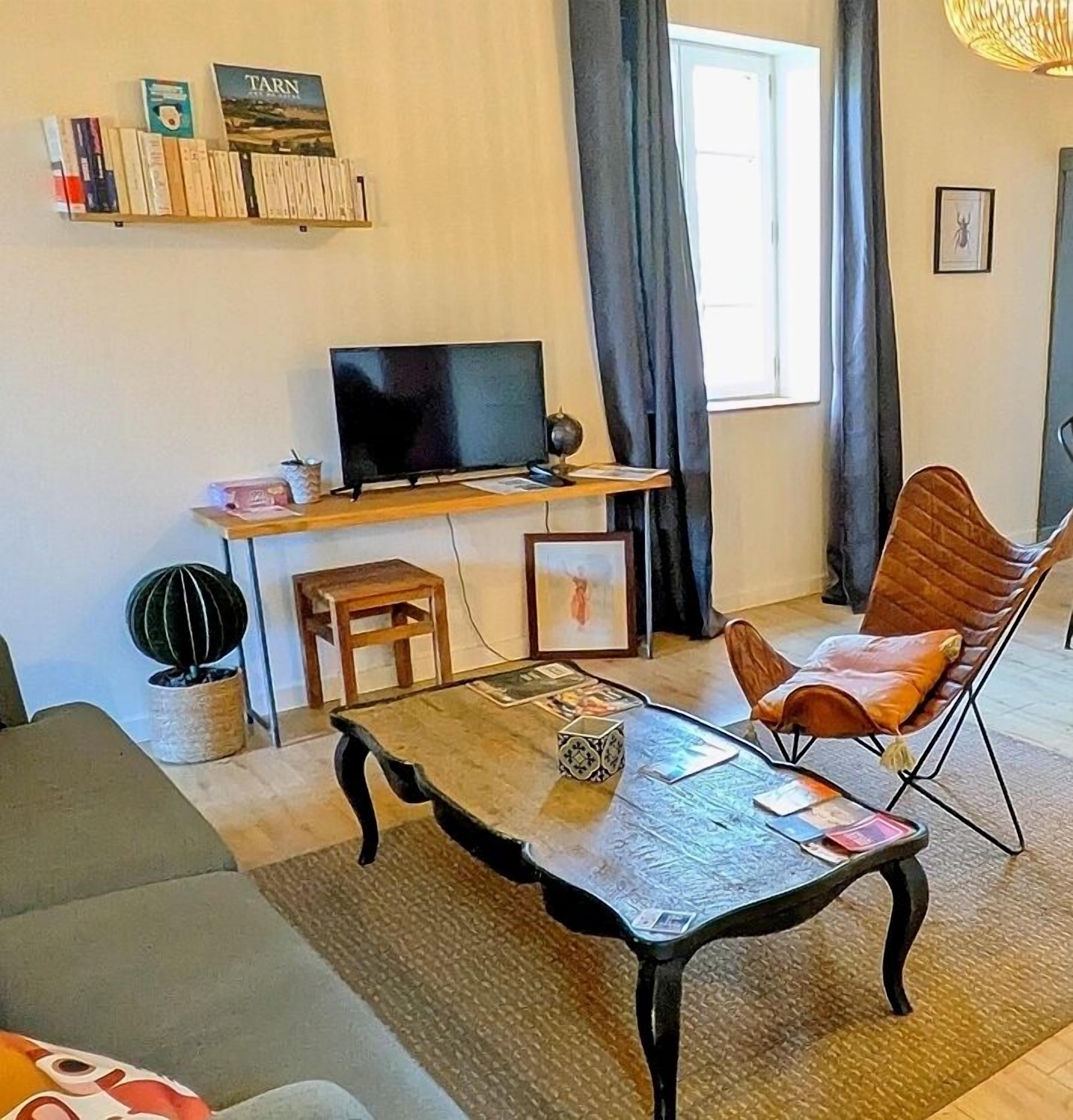 Uma sala de estar com sofá, mesa de centro, televisão, estante de livros e janela com cortinas.