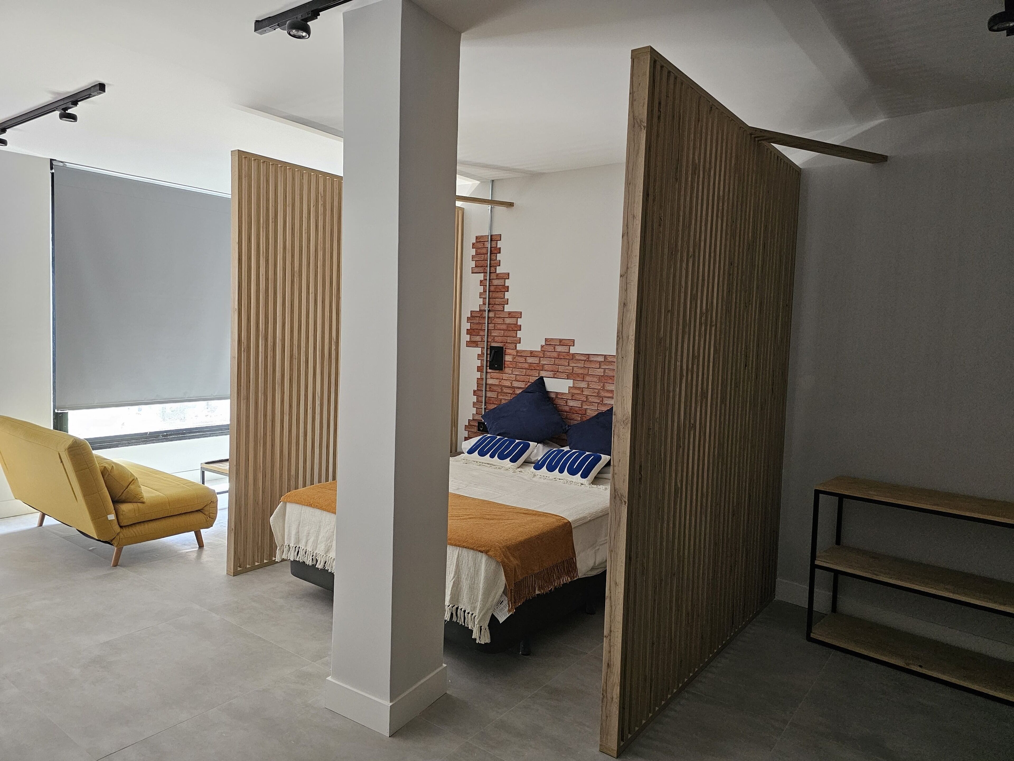 Quarto moderno com cama, poltrona amarela, divisória de madeira e parede de tijolos como destaque.