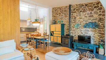 Uma sala de estar aconchegante com uma parede de pedra, lareira e televisão. Há uma área de jantar com mobiliário de madeira e uma cozinha ao fundo.