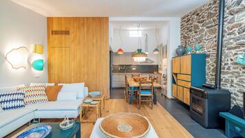 Uma sala de estar moderna com sofá, mesa de centro e área de refeições com cadeiras e mesa. Uma cozinha com fogão e armários é visível ao fundo.