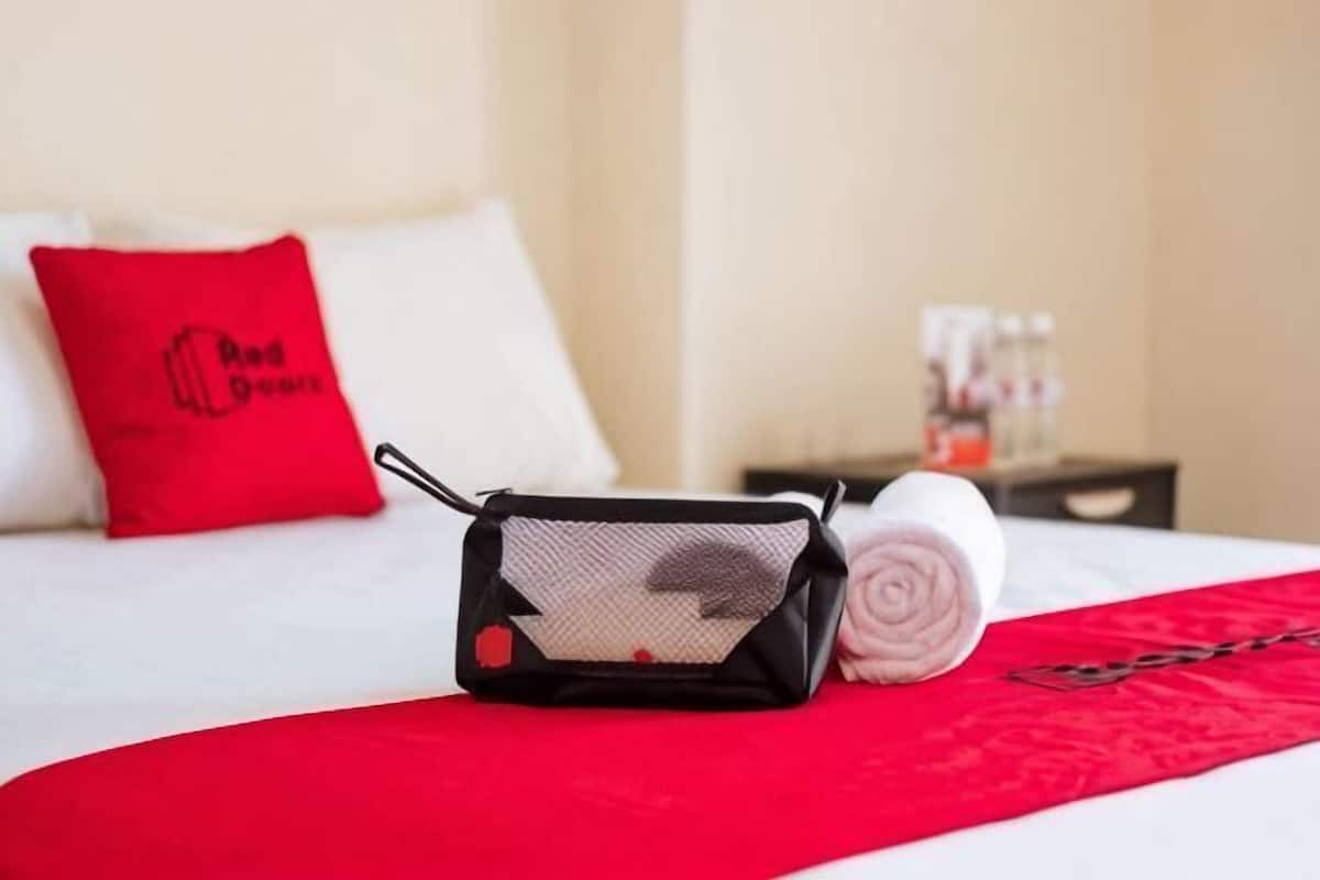 Quarto de hotel com uma cama, um cobertor vermelho, uma toalha enrolada, um travesseiro vermelho e um pequeno objeto preto sobre a cama.