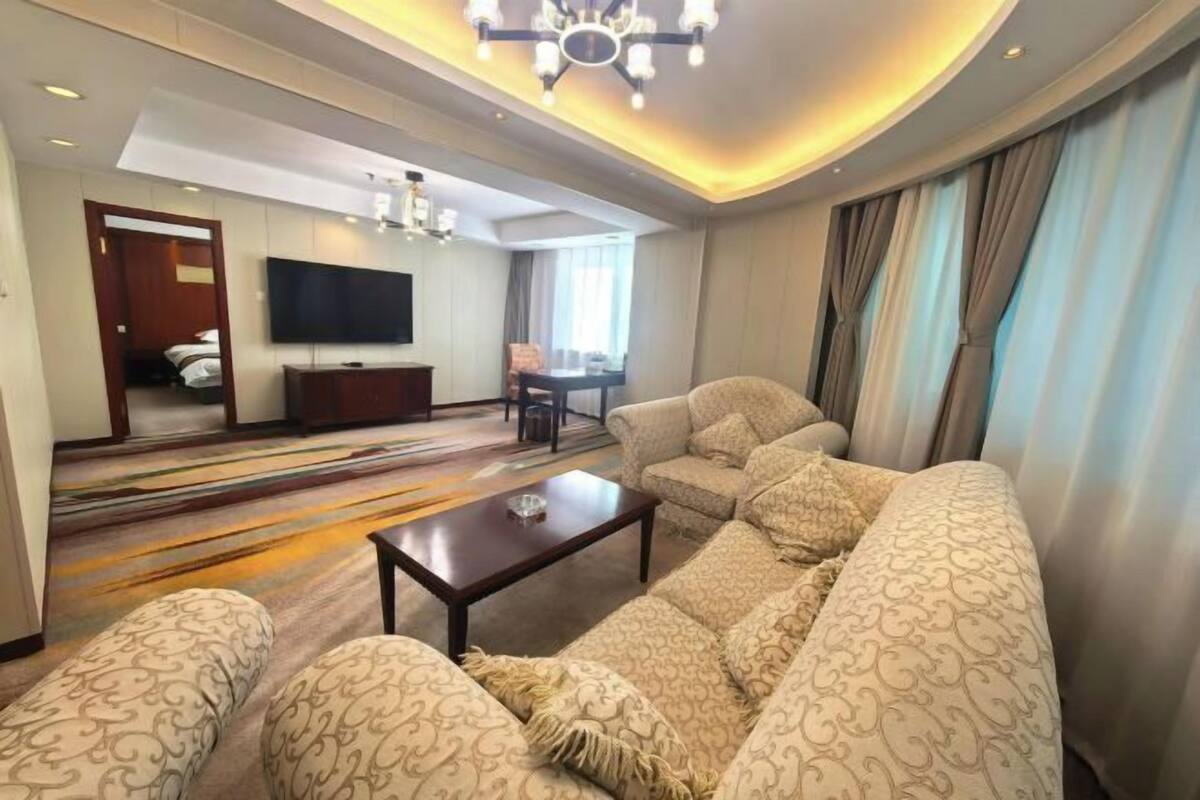 Quarto de hotel com um sofá grande, uma televisão, uma mesa de jantar e um lustre.
