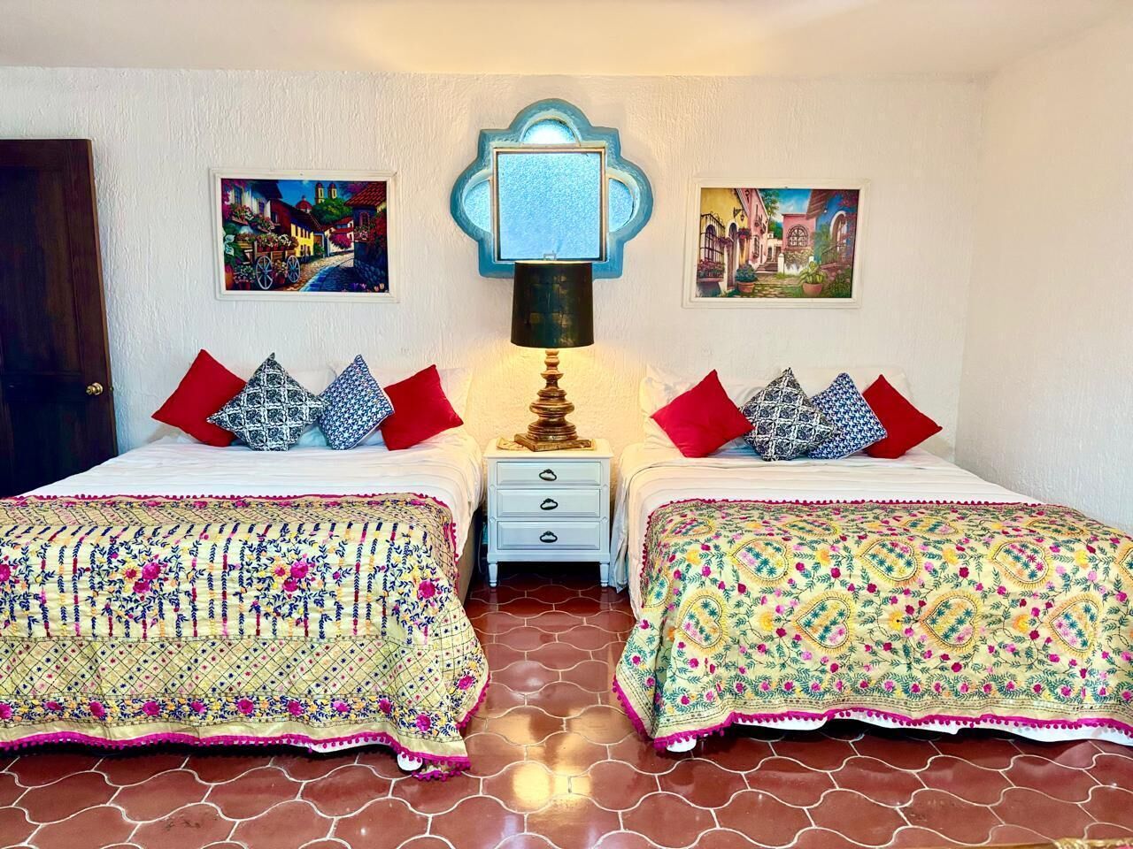 Quarto com duas camas, cada uma com colchas estampadas e almofadas vermelhas. Há uma mesa de cabeceira com abajur e uma gavetinha entre as camas. As paredes são decoradas com quadros emoldurados.