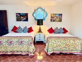 Quarto com duas camas, cada uma com colchas estampadas e almofadas vermelhas. Há uma mesa de cabeceira com abajur e uma gavetinha entre as camas. As paredes são decoradas com quadros emoldurados.