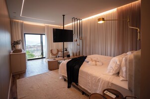 Quarto de hotel moderno com uma cama grande, televisão, mesa de cabeceira, abajur e vista para o exterior.