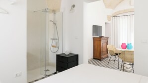 Casa de banho moderna com chuveiro envolto em vidro, mesa de cabeceira preta e área de refeições com cadeiras de vime e uma mesa.