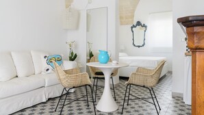 Um sofá branco com uma almofada colorida, duas cadeiras de vime, uma mesa branca com um jarro azul, um espelho e um armário de madeira.