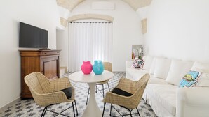 Quarto com sofá branco, cadeiras de vime, uma pequena mesa com dois vasos, TV na parede e janela com cortinas.