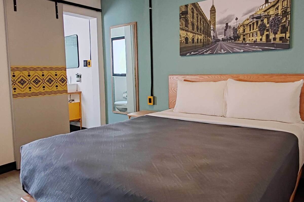 Quarto com cama, espelho, porta e pintura de paisagem urbana na parede.