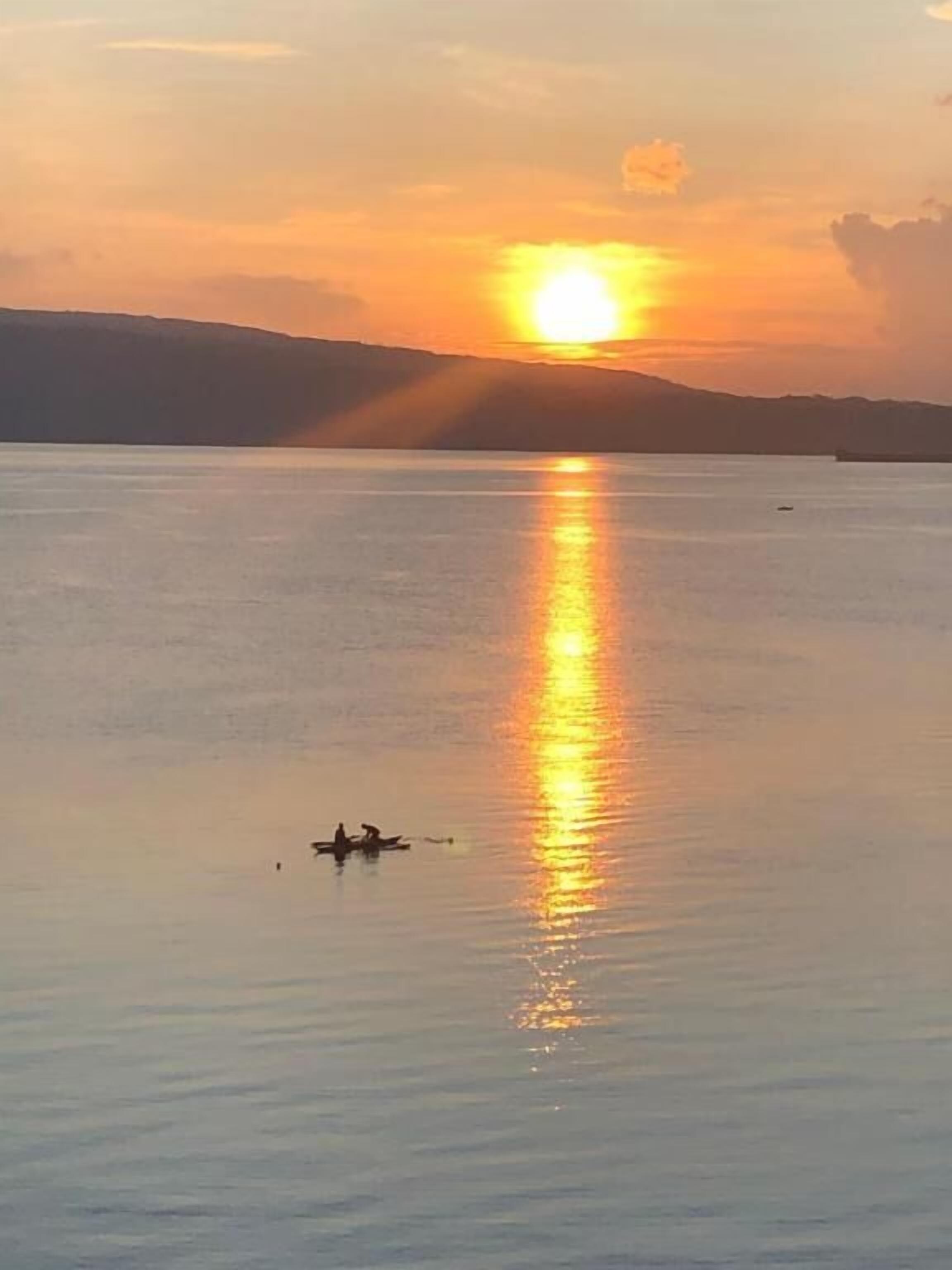 Água calma refletindo o sol ao pôr do sol, com a silhueta de um barco e montanhas ao longe.