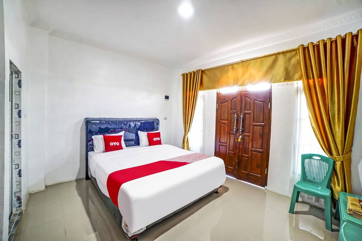 Quarto de hotel com uma cama, porta de madeira, uma cadeira verde e uma pequena mesa.