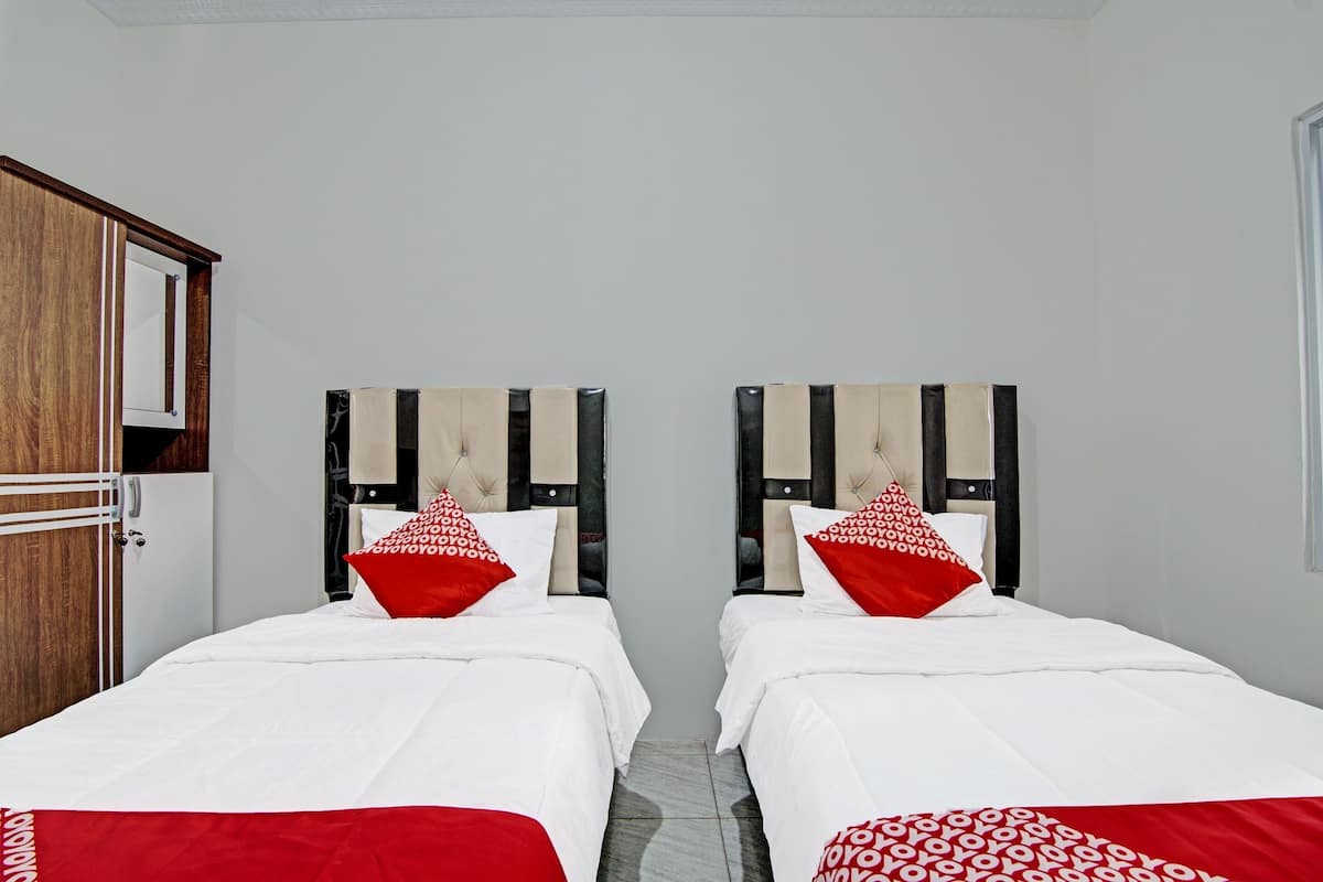 Quarto de hotel com duas camas, cada uma com colcha e almofadas estampadas em vermelho e branco. O quarto possui um guarda-roupa de madeira e uma porta branca.