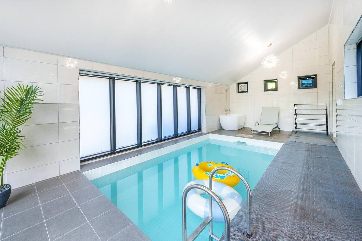 Piscina interna com parede de vidro, planta em vaso, boia inflável amarela e azul, e banheira branca.