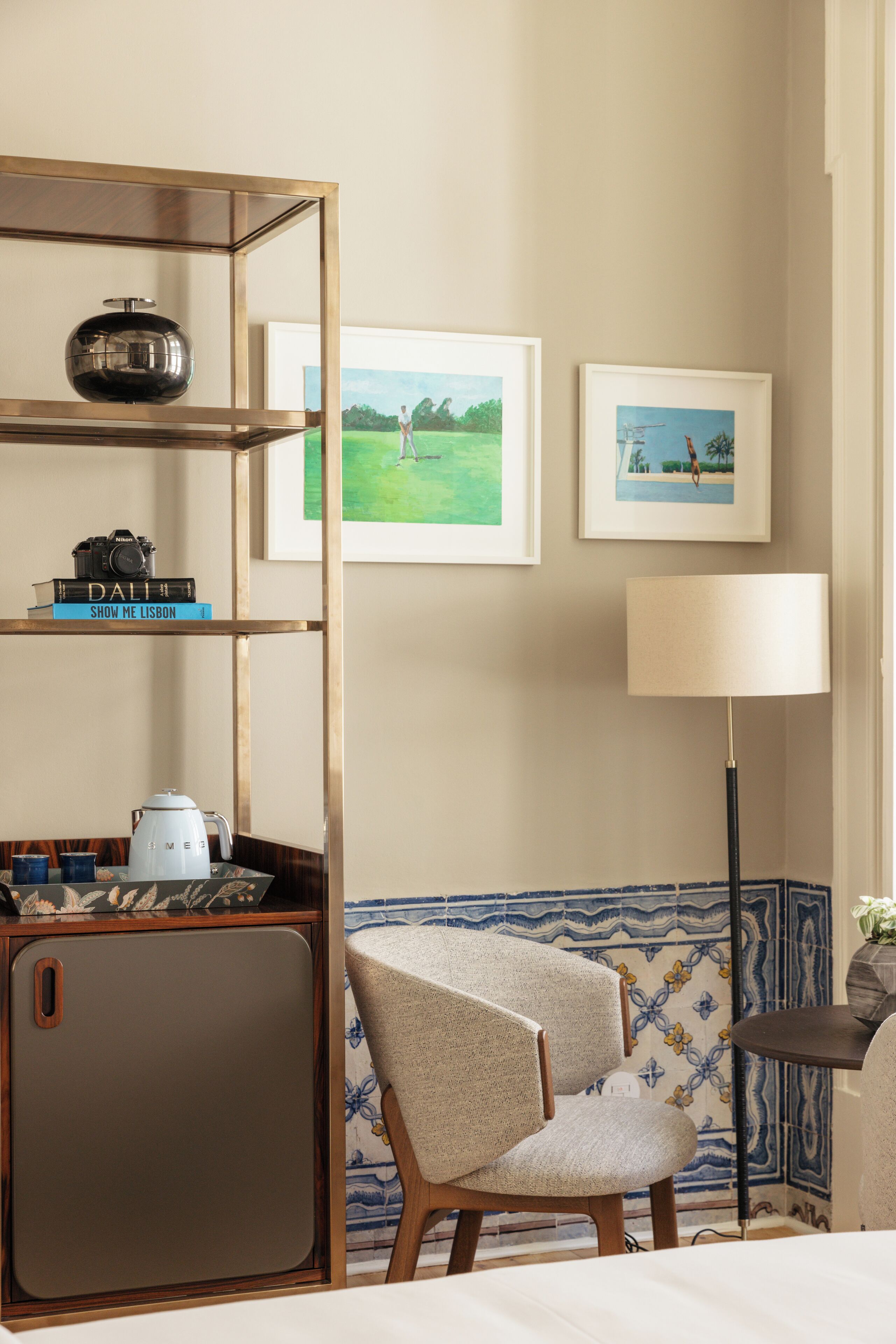 Quarto de hotel com estante de madeira, uma mesa com luminária, duas cadeiras, uma cama e uma parede com quadros.