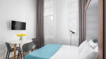 Quarto de hotel com cama, escrivaninha com cadeira, televisão fixada na parede e janela com cortinas.