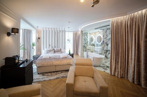 Quarto moderno com uma cama grande, área de estar, toucador com painel de mármore e uma janela ampla com cortinas.
