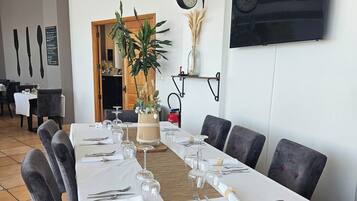Uma área de jantar com uma mesa comprida posta para seis pessoas, cercada por cadeiras. Há uma planta em vaso e um relógio fixado na parede.
