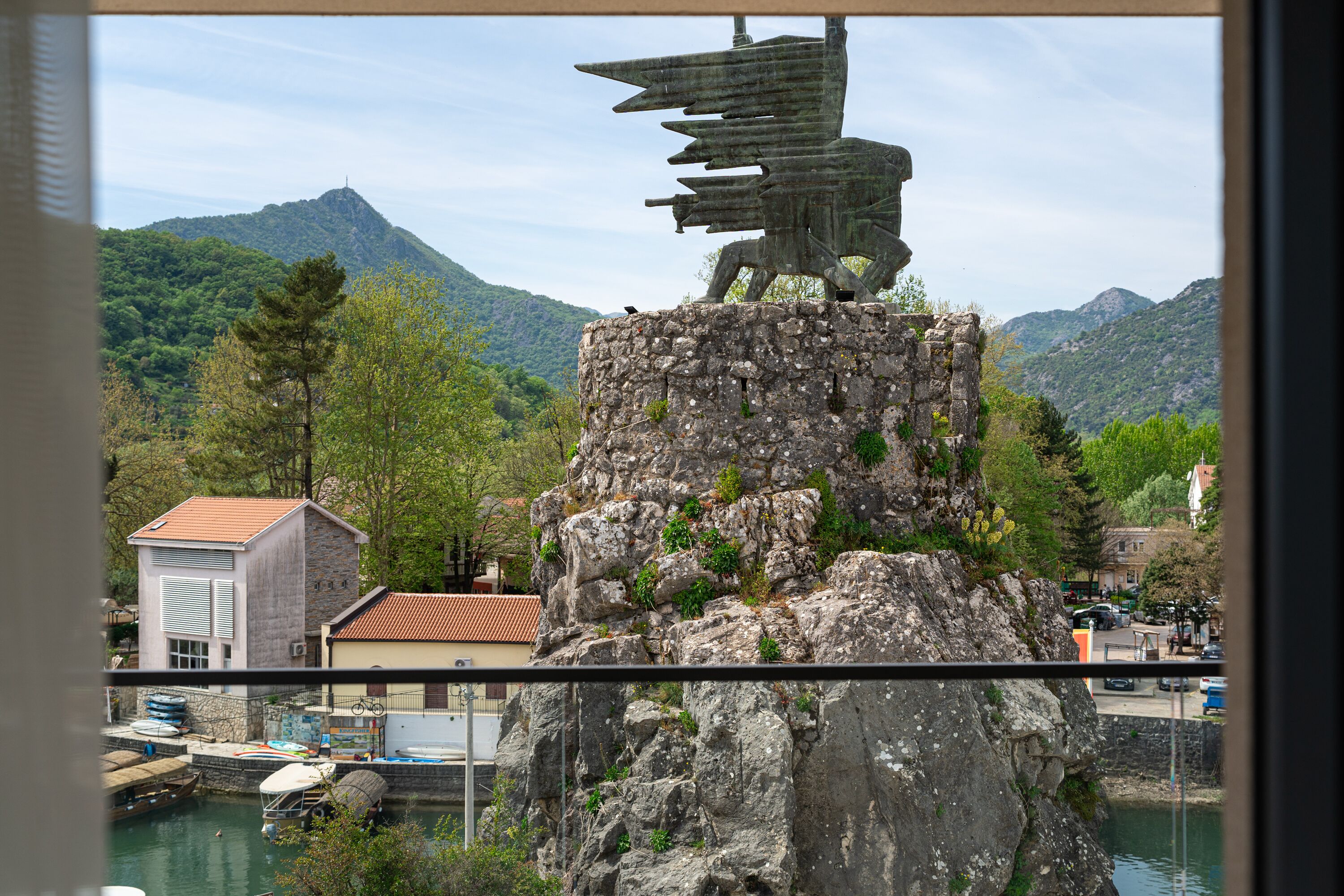 Um afloramento rochoso com uma grande escultura de pássaro no topo, com vista para um corpo de água com barcos e edifícios ao fundo.
