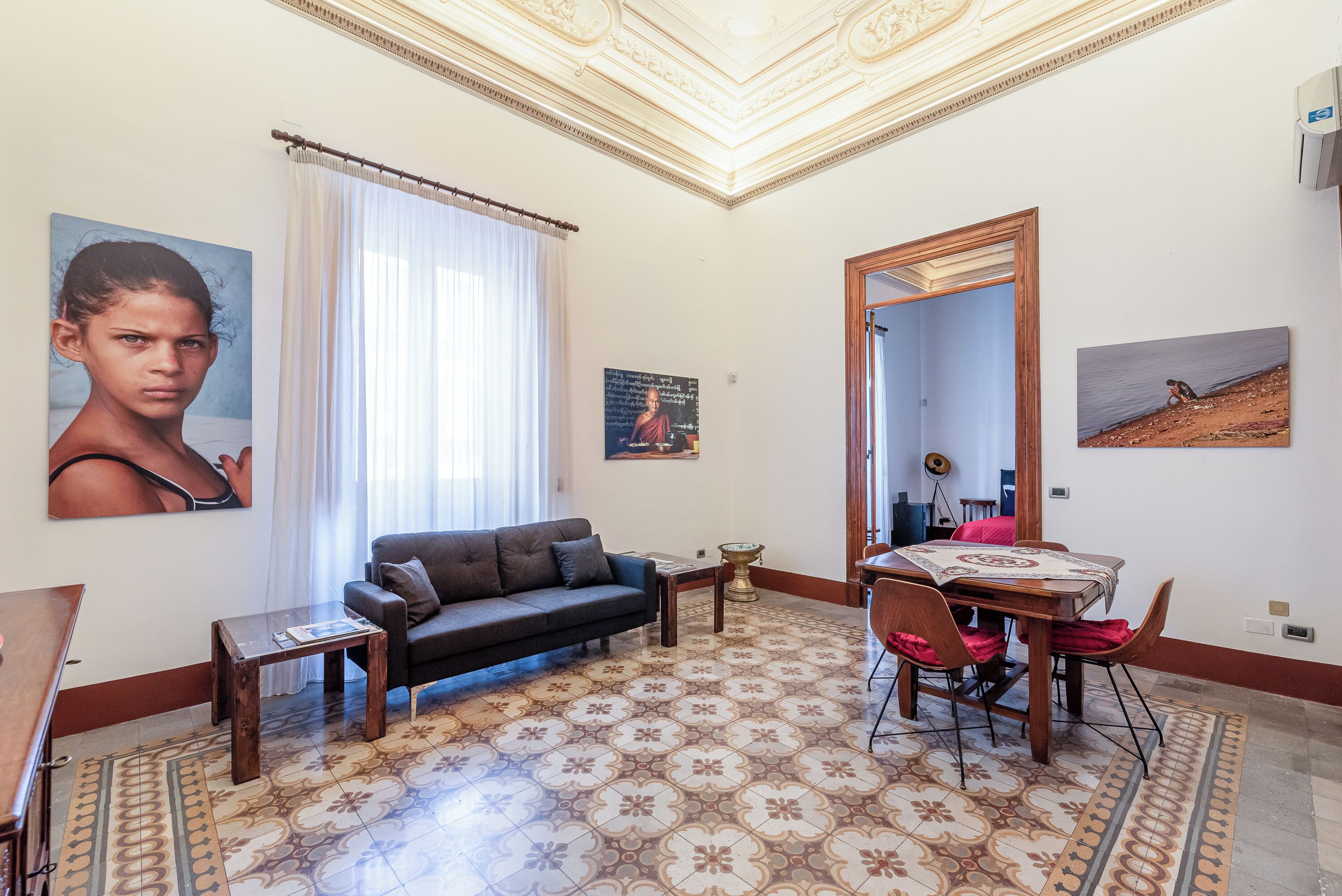 Sala de estar com sofá, mesa de centro e mesa de jantar com cadeiras. Há azulejos decorativos no chão e um quadro na parede.