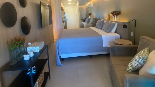 Quarto de hotel moderno com cama, sofá, mesa pequena e console com objetos decorativos.