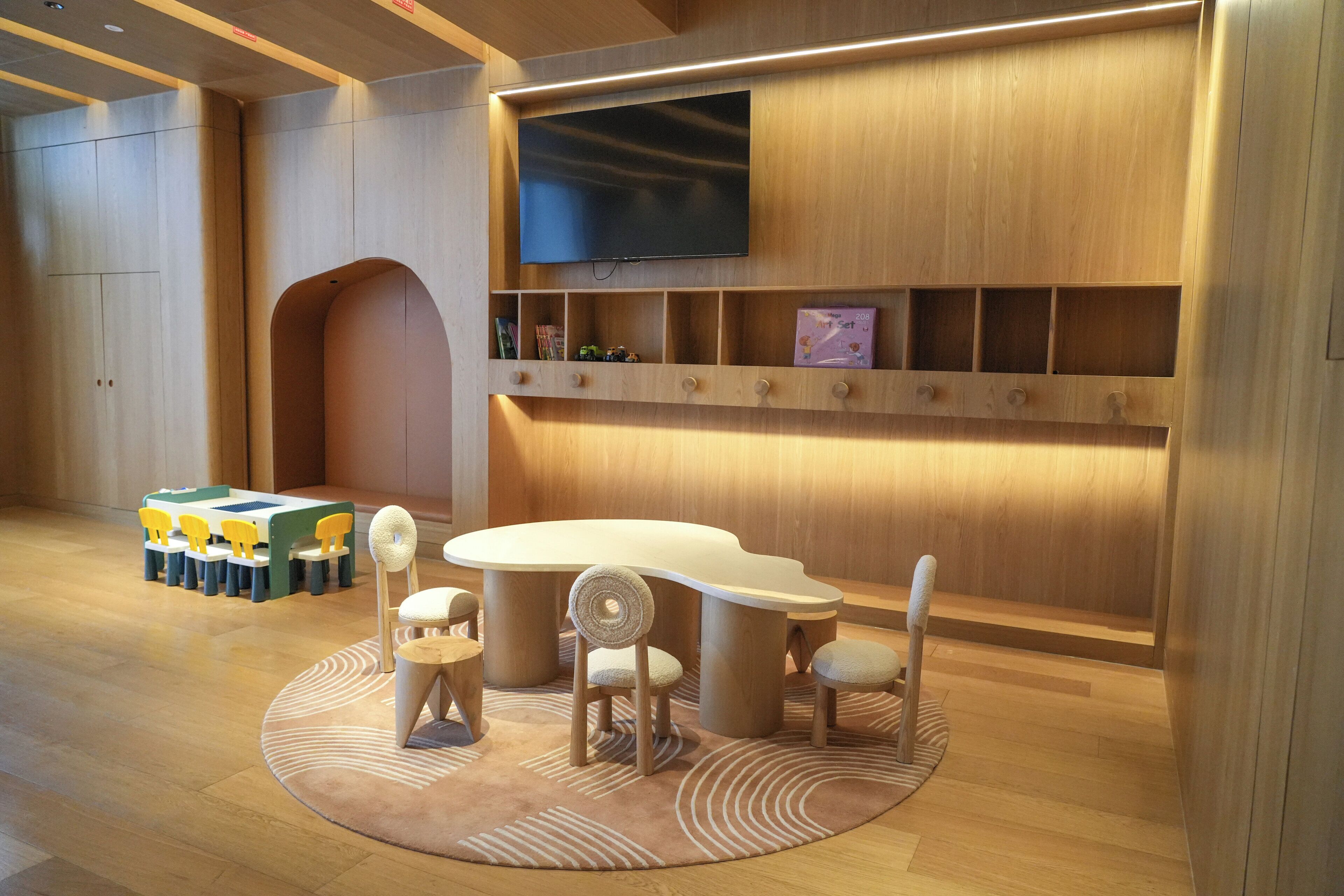 Quarto moderno com piso de madeira, uma mesa redonda com cadeiras, televisão fixada na parede e uma estante com diversos objetos.