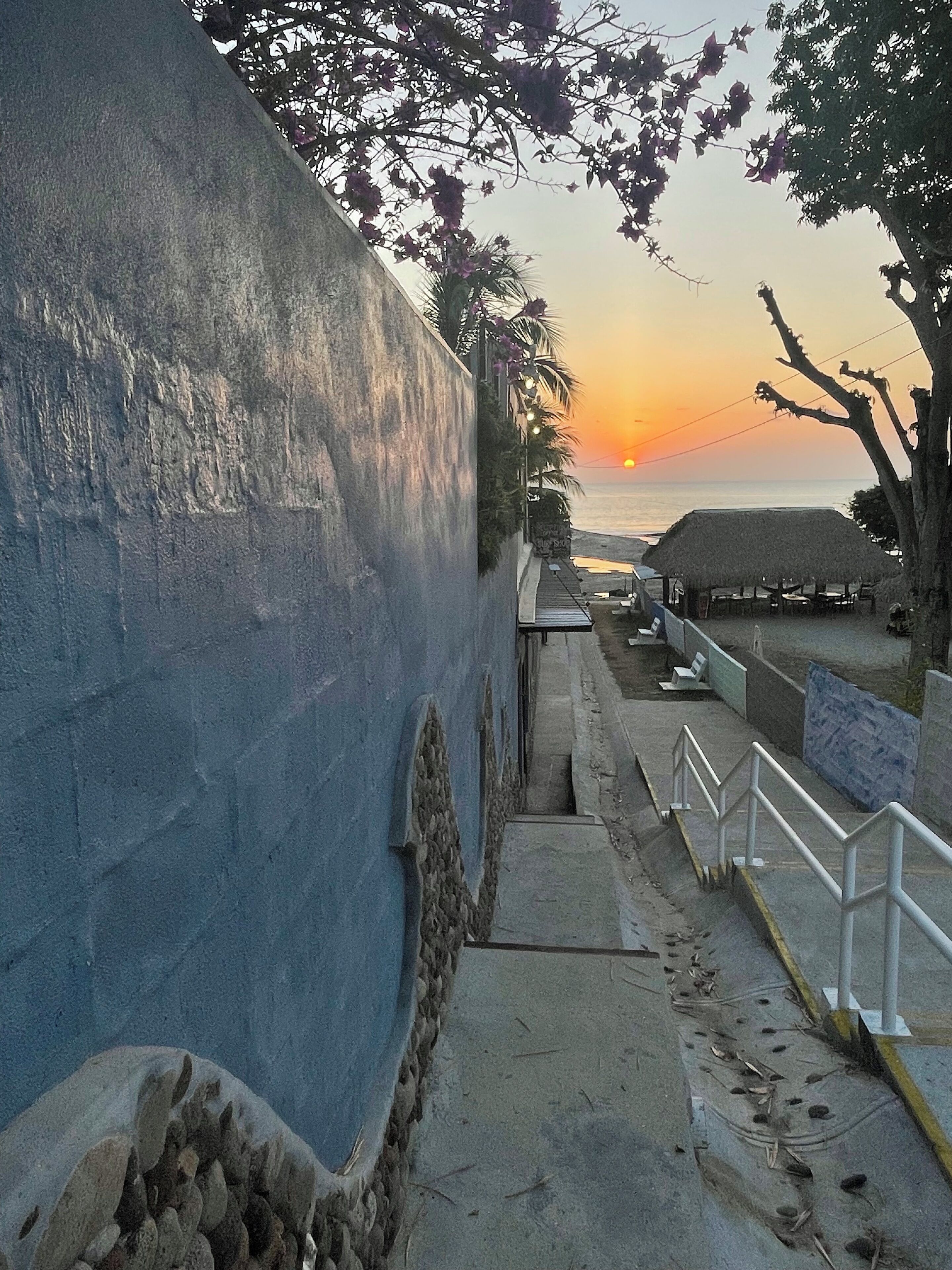 Uma passarela costeira com vista para o pôr do sol sobre o mar, um edifício com telhado de palha e um caminho pavimentado com grades.