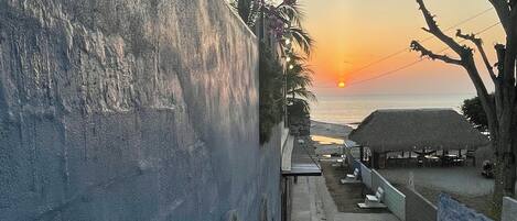 Uma passarela costeira com vista para o pôr do sol sobre o mar, um edifício com telhado de palha e um caminho pavimentado com grades.