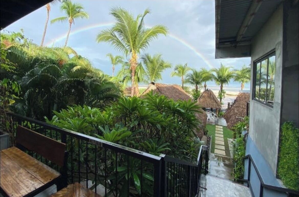 Varanda com vista para um jardim tropical, palmeiras e uma praia.