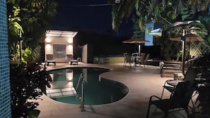 Área de piscina à noite com espreguiçadeiras e guarda-sóis, cercada por vegetação.