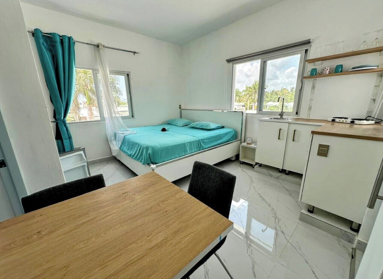 Quarto compacto com cama, pequeno espaço de cozinha, mesa de jantar e uma janela.