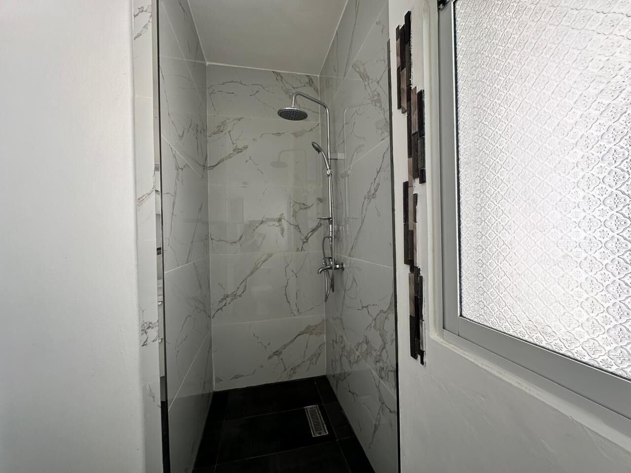 Um banheiro pequeno e moderno, com chuveiro, azulejos de mármore e uma janela de vidro fosco.