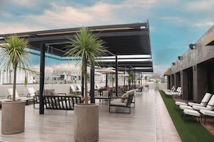 Um lounge no terraço com mesas ao ar livre, palmeiras em vasos e vista para a cidade.