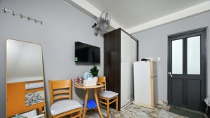 Quarto compacto com cama, televisão na parede, mesinha com cadeiras, geladeira e porta com janela.