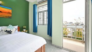 Quarto com cama, janela com cortinas azuis, quadro na parede e varanda com guarda-corpo.