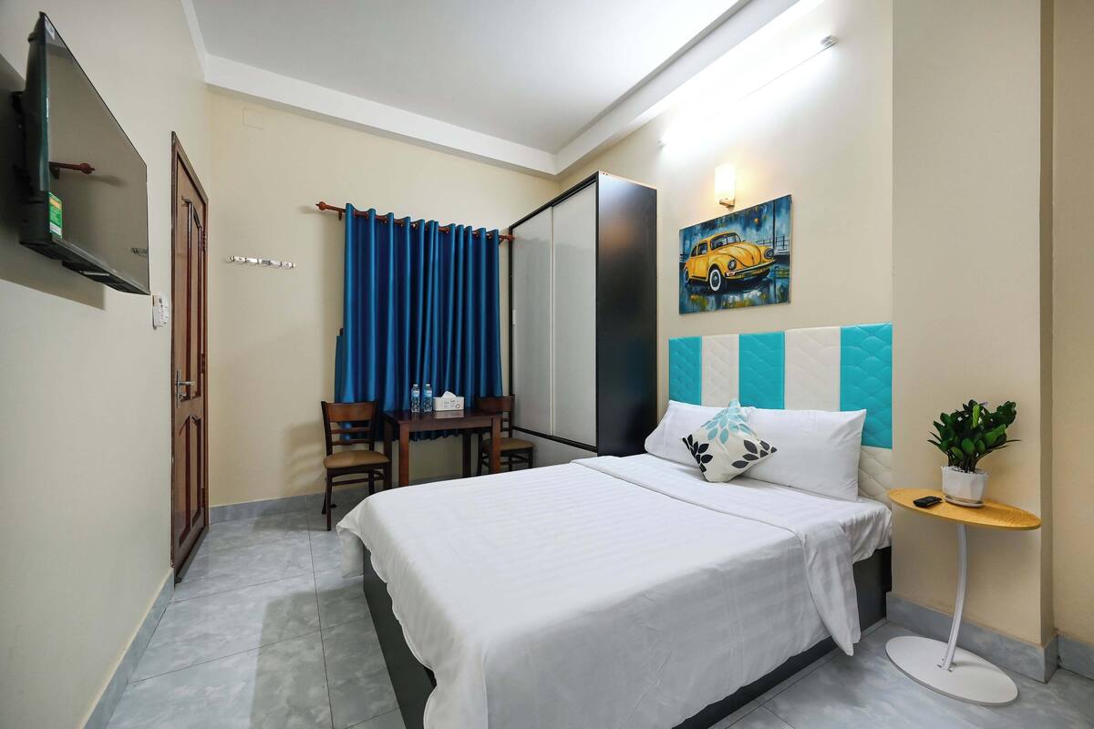 Quarto de hotel com cama, escrivaninha, cadeira, televisão e um quadro na parede.