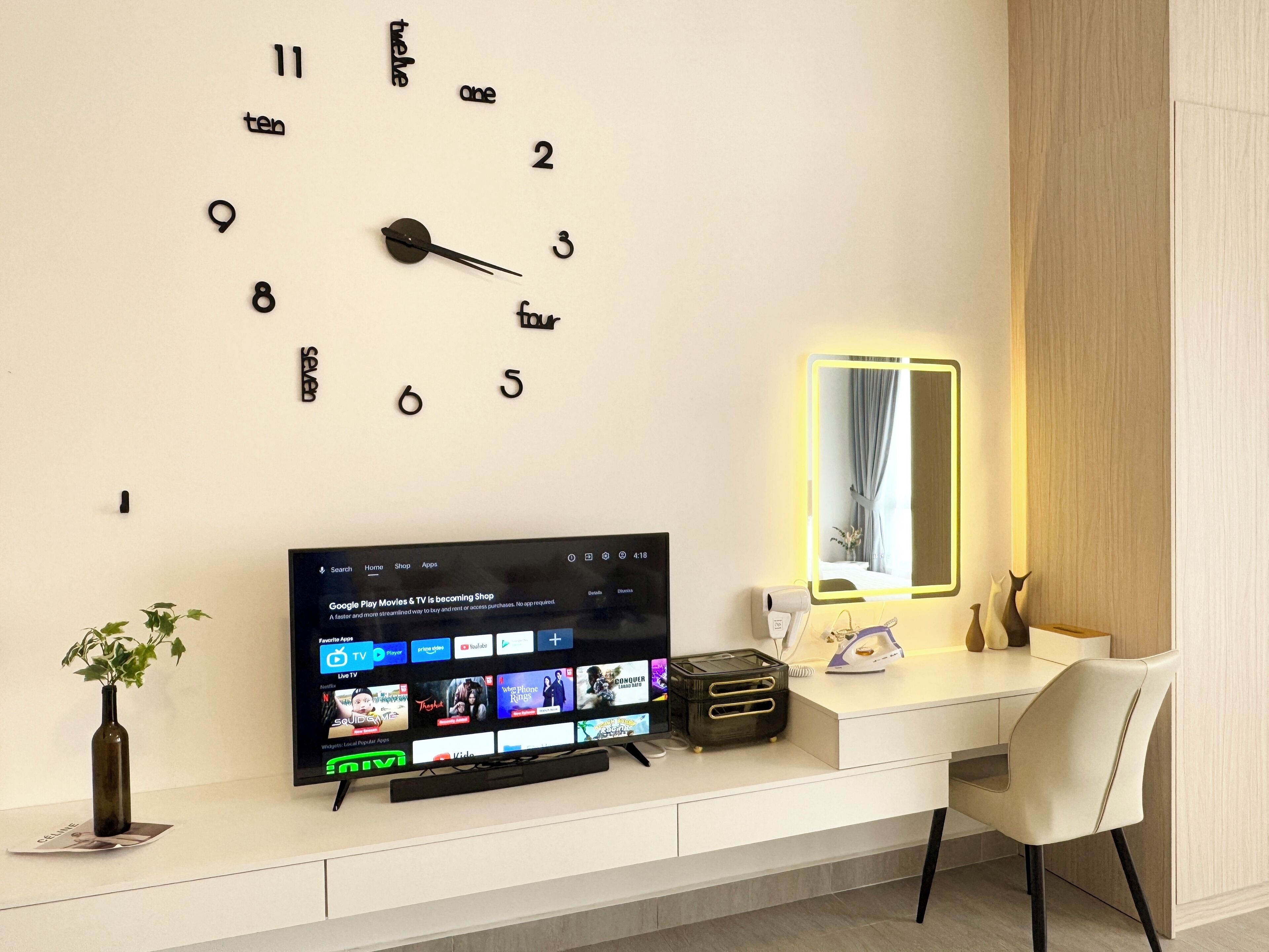 Quarto moderno com relógio de parede, TV de tela plana exibindo um serviço de streaming, escrivaninha com cadeira e espelho iluminado.