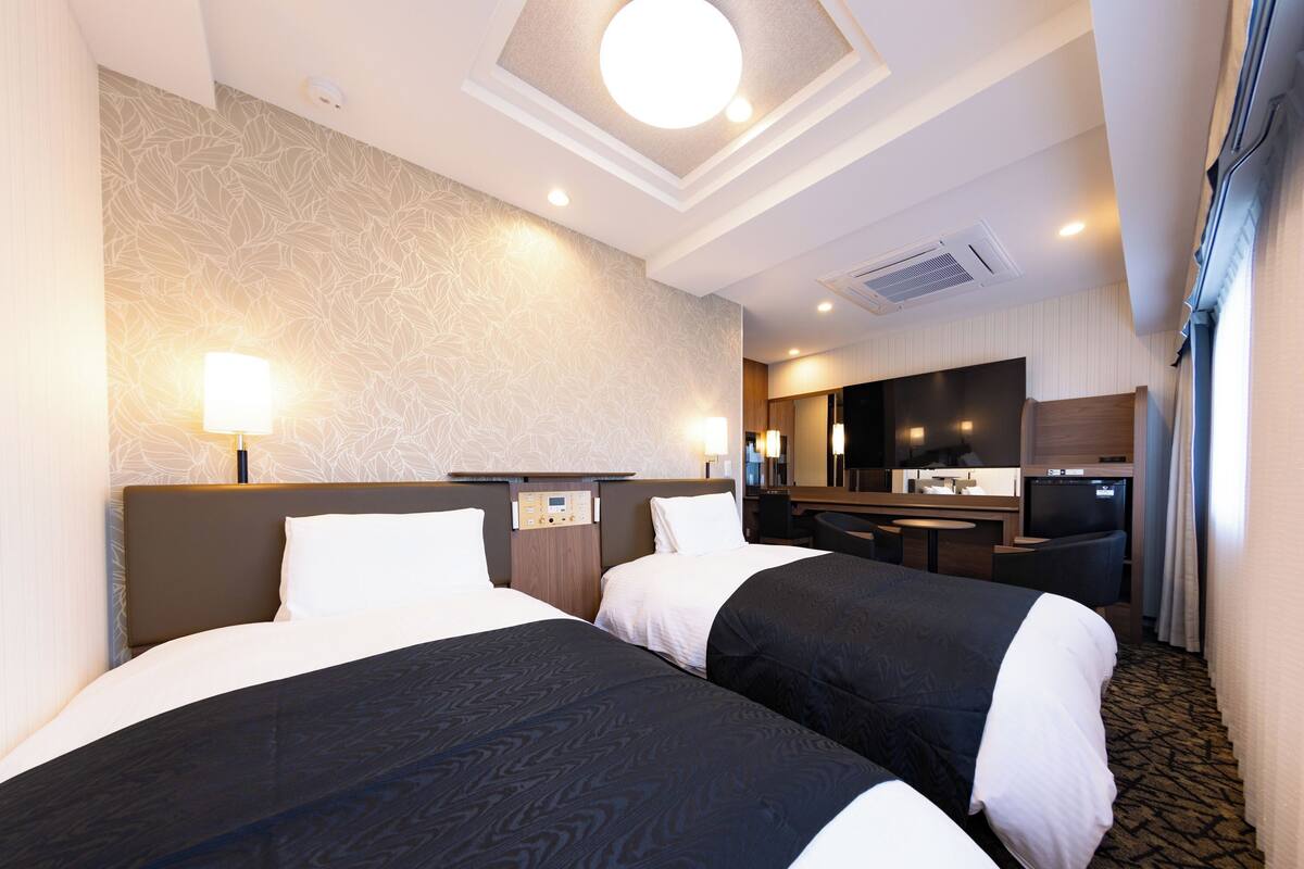 Quarto de hotel com duas camas, uma escrivaninha, uma cadeira, uma mesinha, um espelho e uma cozinha compacta.