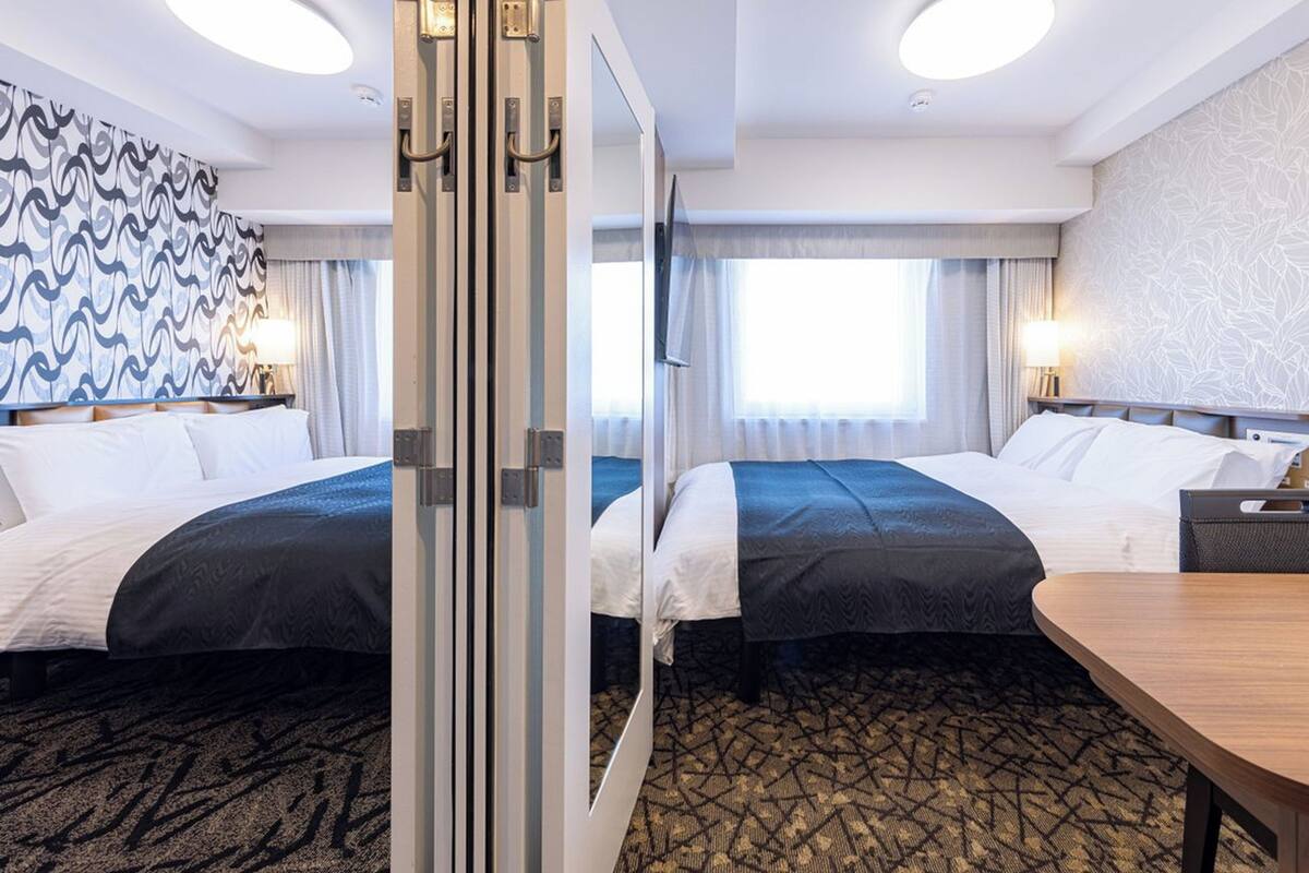 Quarto de hotel com duas camas, uma mesa de madeira, carpete estampado e uma janela grande com cortinas.