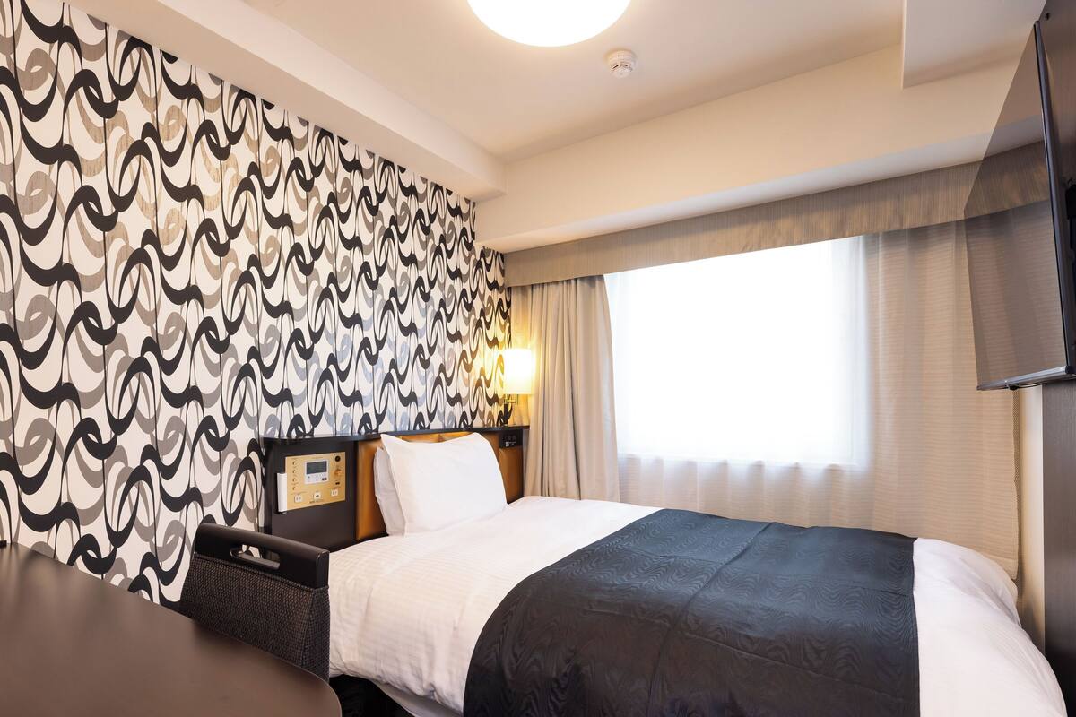 Quarto de hotel com cama, escrivaninha, parede com papel de parede estampado, janela com cortinas e TV fixada na parede.