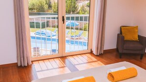 Quarto com uma grande porta de vidro deslizante que dá para uma área de piscina, uma cama com duas toalhas enroladas, uma poltrona cinza com almofada amarela e piso de madeira.