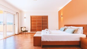 Quarto com cama grande, guarda-roupa de madeira, uma cadeira e porta de vidro deslizante para área externa.