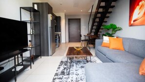 Uma sala de estar moderna com um sofá cinzento, um móvel para entretenimento preto e uma mesa de centro. Uma escadaria é visível ao fundo, levando a uma área de refeições.