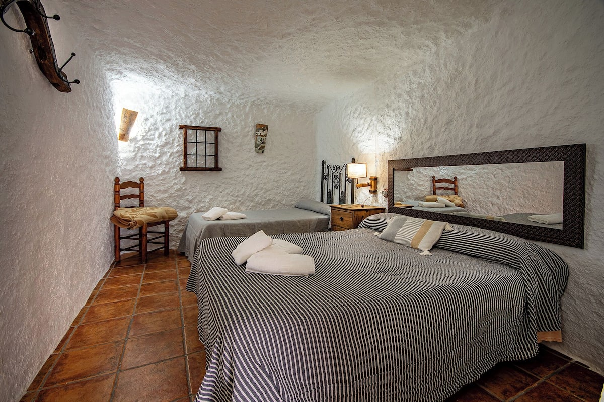 Quarto com duas camas, uma cadeira de madeira, uma mesa de cabeceira, um espelho e uma janela.