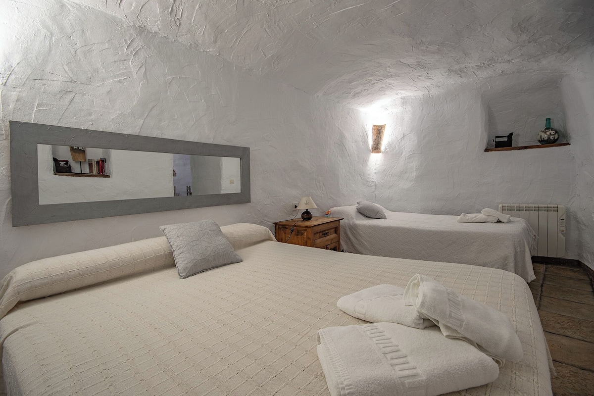 Quarto com duas camas, uma mesinha de cabeceira, um espelho e uma estante com objetos.