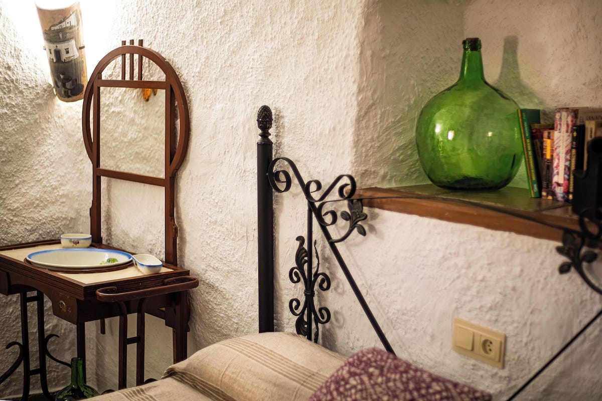Quarto rústico com uma cadeira de madeira, uma mesa pequena com uma tigela, uma garrafa verde em uma prateleira e um luminário fixado na parede.