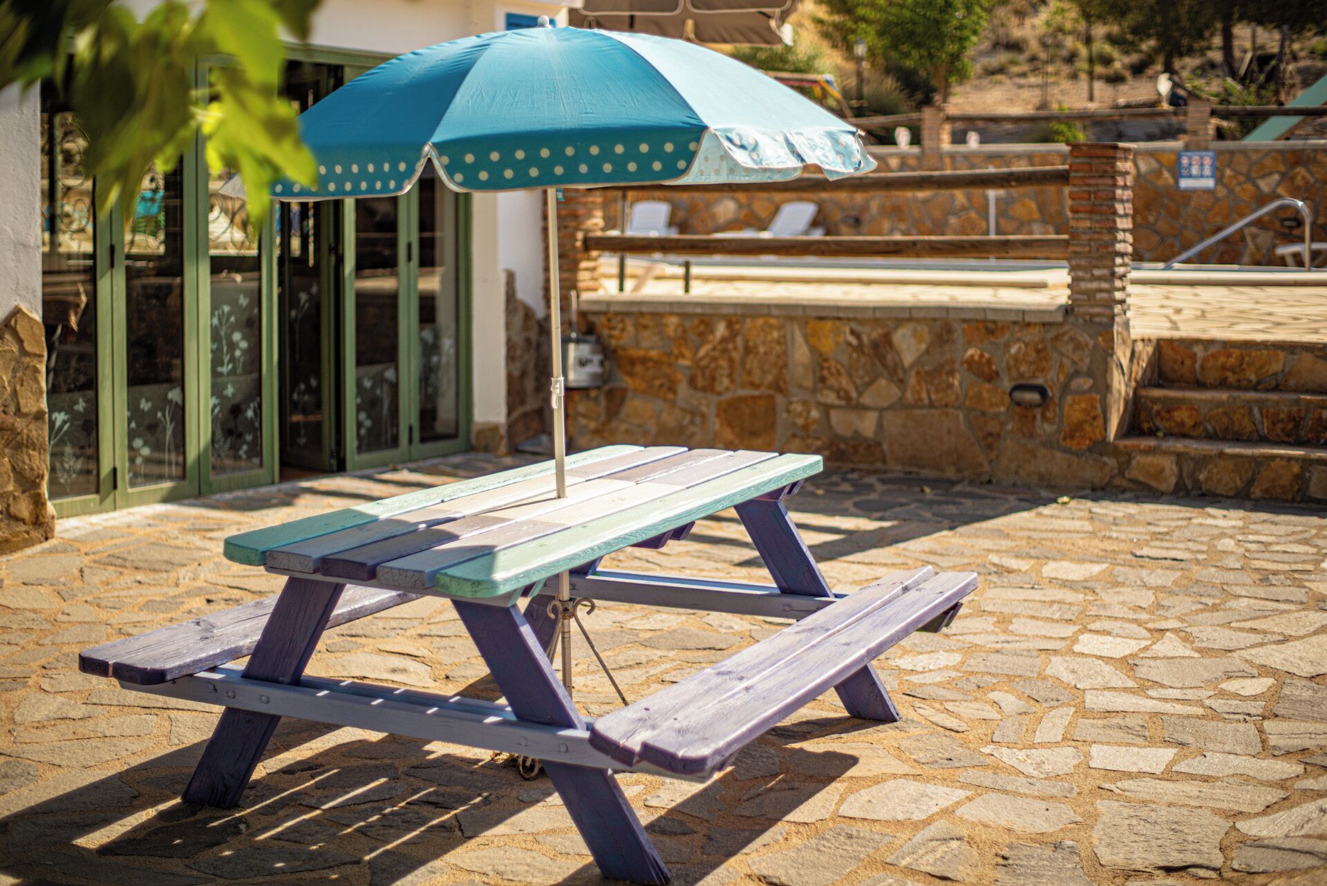 Uma mesa de piquenique com um guarda-sol azul, degraus de pedra e um muro de pedra.