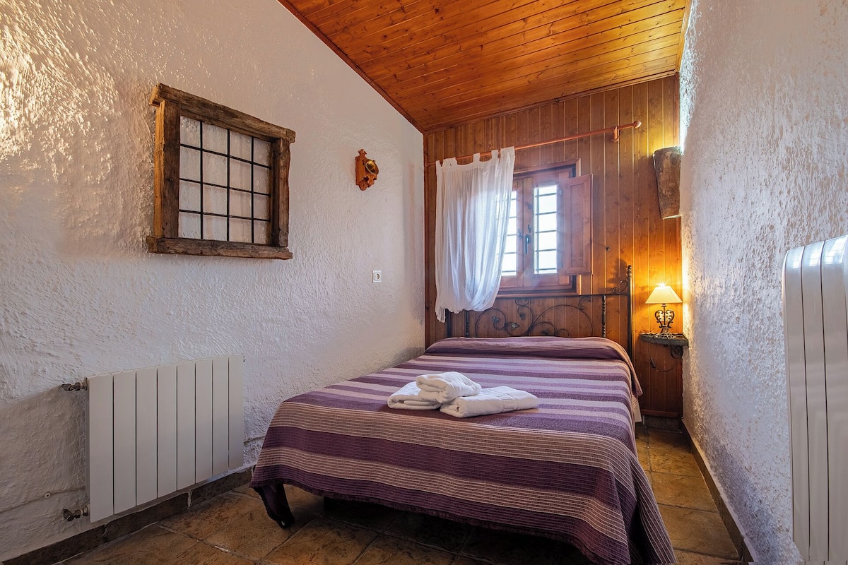 Um quarto aconchegante com teto de madeira, uma cama com roupa de cama listrada, uma janela com moldura de madeira e um abajur.