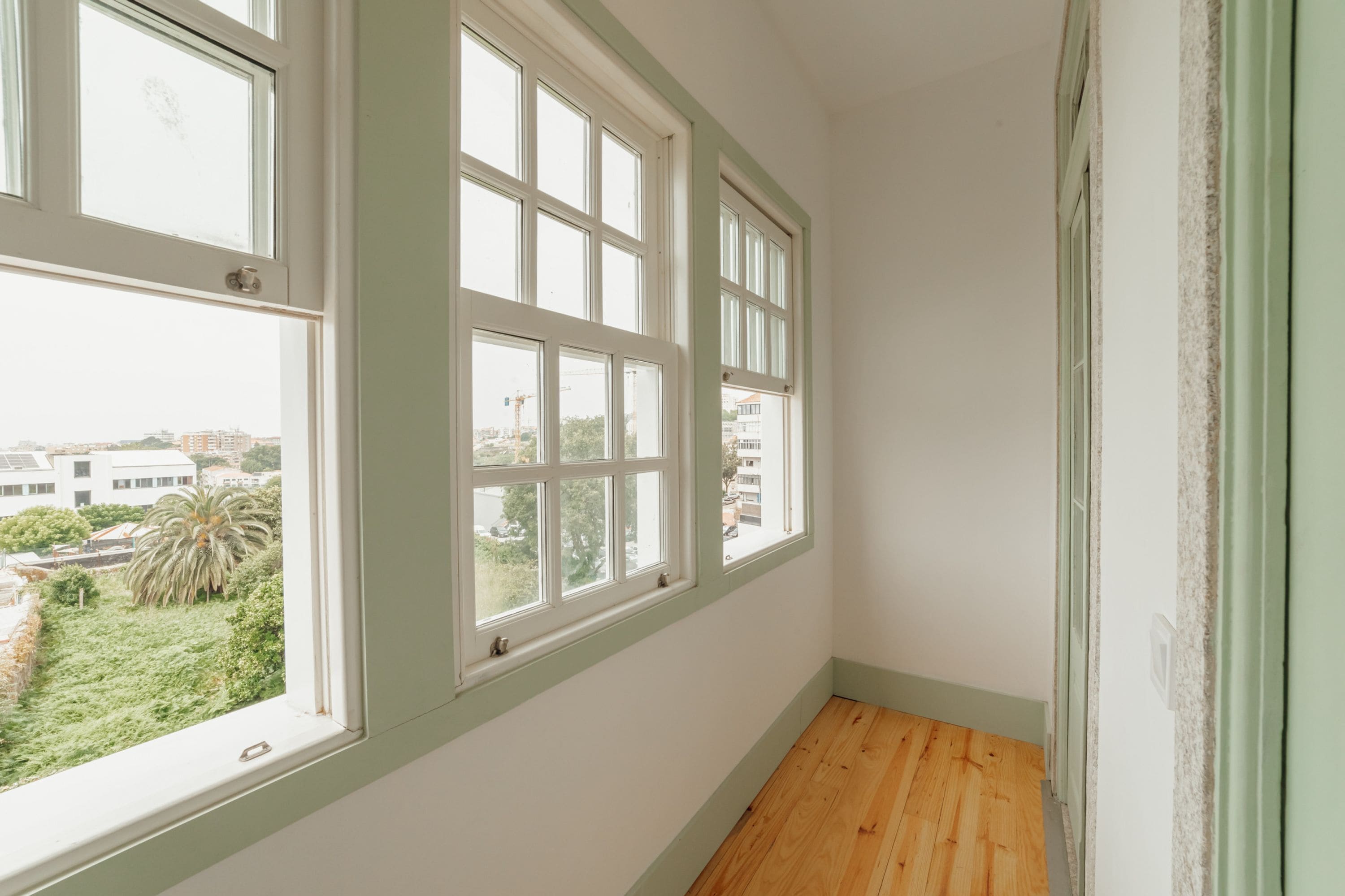 A room with three large windows, wooden flooring, and a view of buildings and greenery outside.