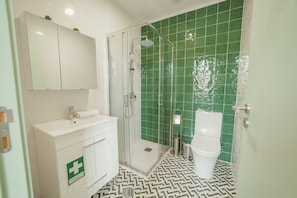 Casa de banho com lavatório branco, cabine de chuveiro em vidro, sanita e paredes revestidas a mosaico verde.